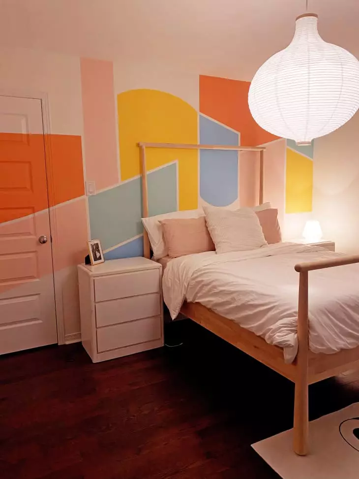 Kamar turu warna-warni kanthi amben kayu IKEA, mural warna-warni geometris sing dicet ing tembok amben lan lawang, lan liontin kertas gantung putih gedhe.