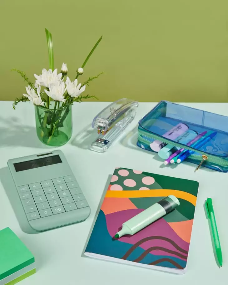 Skrivebord med grønn kalkulator, notatbok, blyantpose og liten vase med blomster