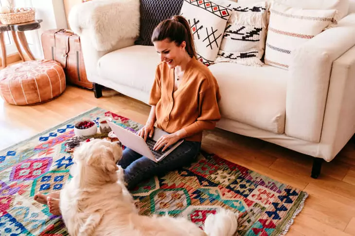 Žena sediaca na podlahe obývačky so psom, pracujúca na notebooku