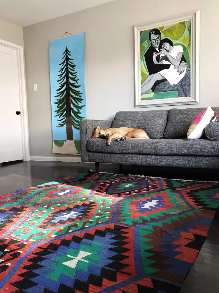 Svetainė su dideliais meno kūriniais, spalvingu kiliminiu kilimėliu ir pilka sofa