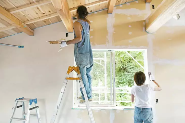 2 ľudia, jeden na rebríku, maľujú steny domu na bielo. Strop je z odhaleného dreva. Je tu okno prepúšťajúce denné svetlo a vonku sú stromy