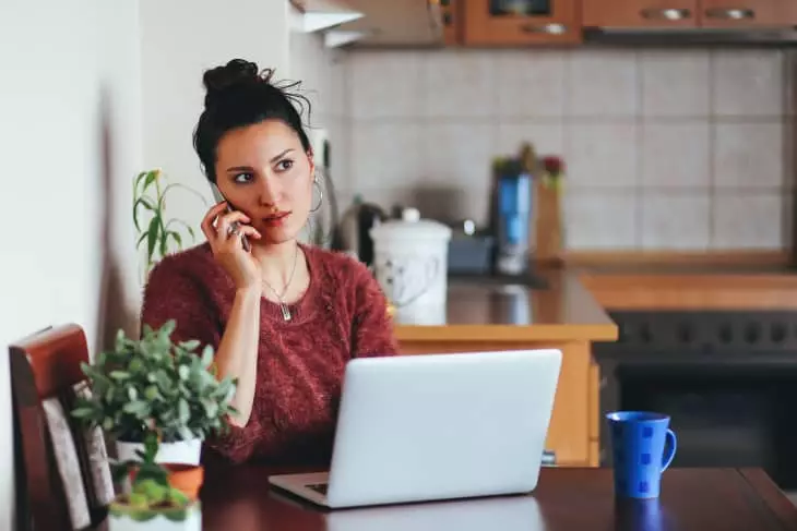 mujer sentada en la cocina con una computadora portátil. Ella está al teléfono y tiene una taza de café azul.